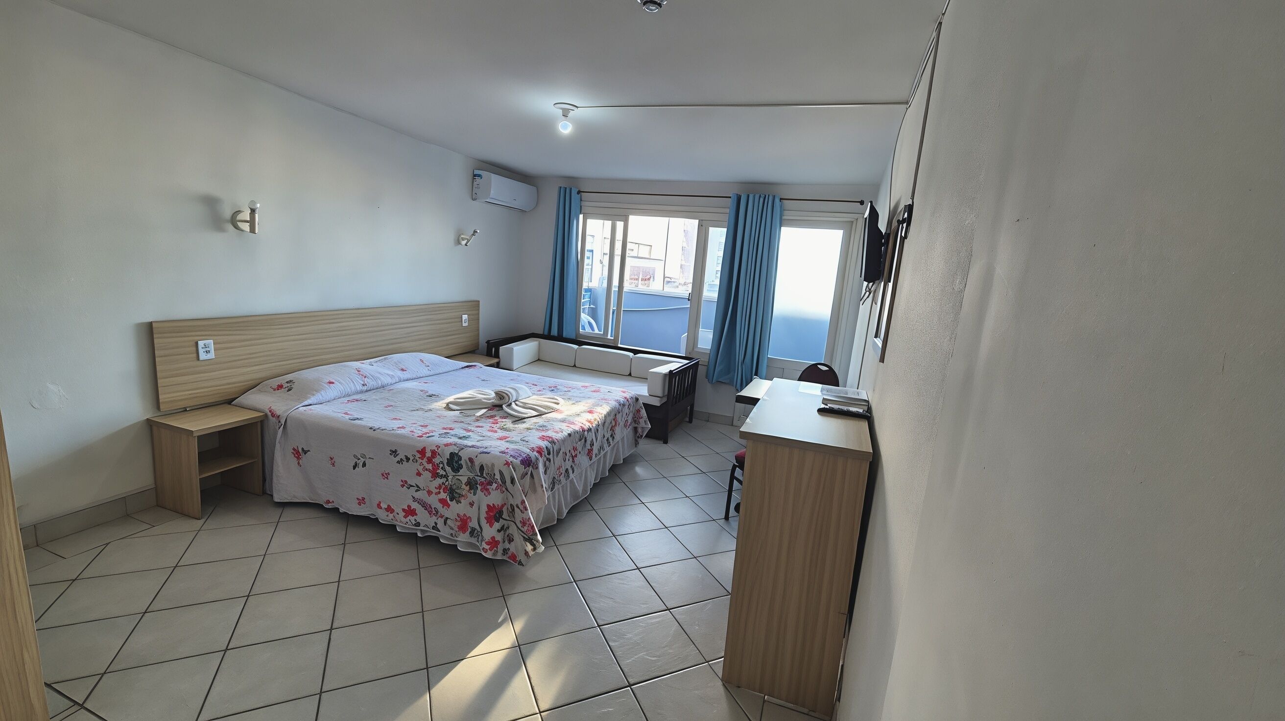 Quarto com cama, mesa de cabeceira, escrivaninha, cadeira e janela com cortinas.