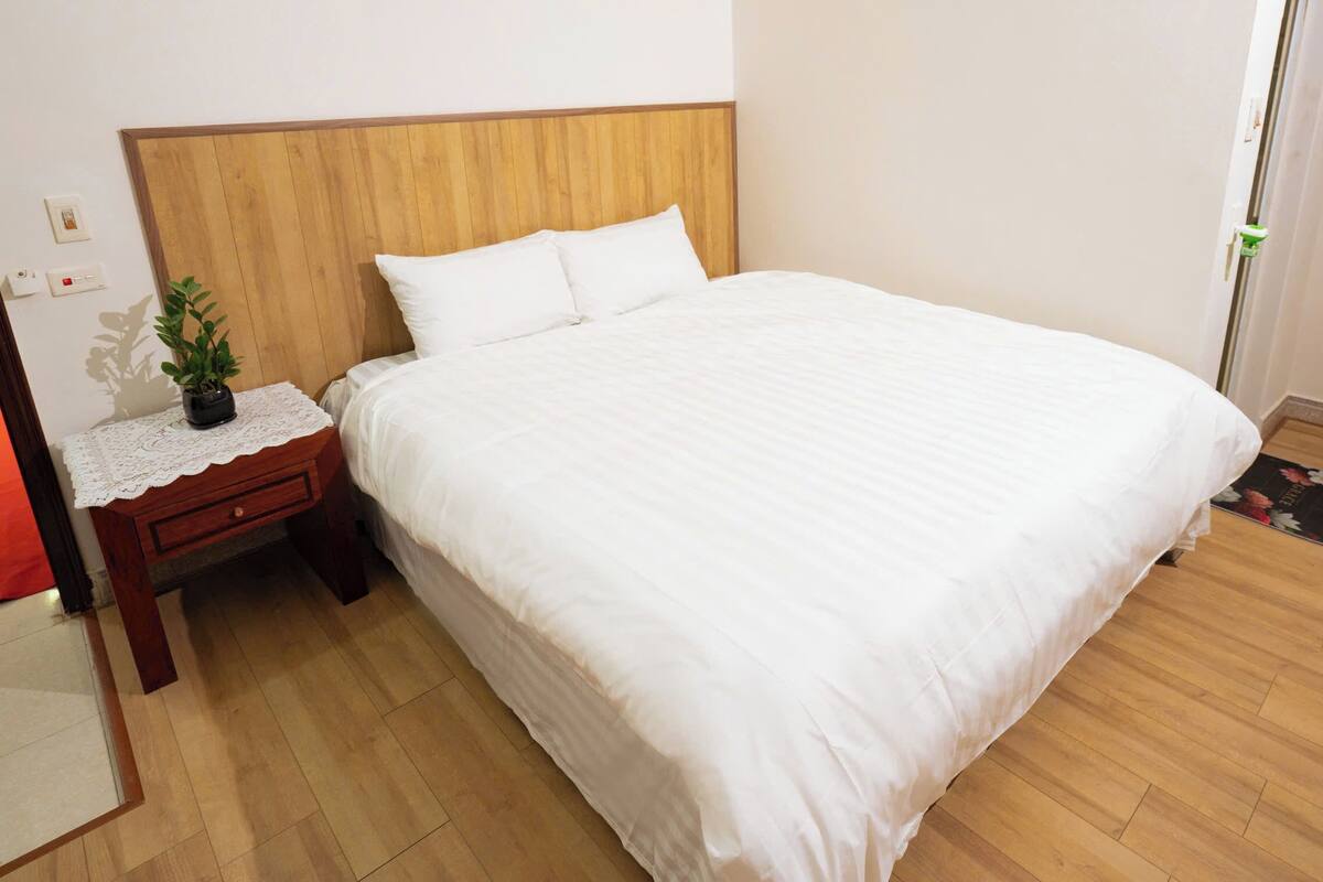Cama bem arrumada com roupa de cama branca em quarto com piso e cabeceira de madeira.