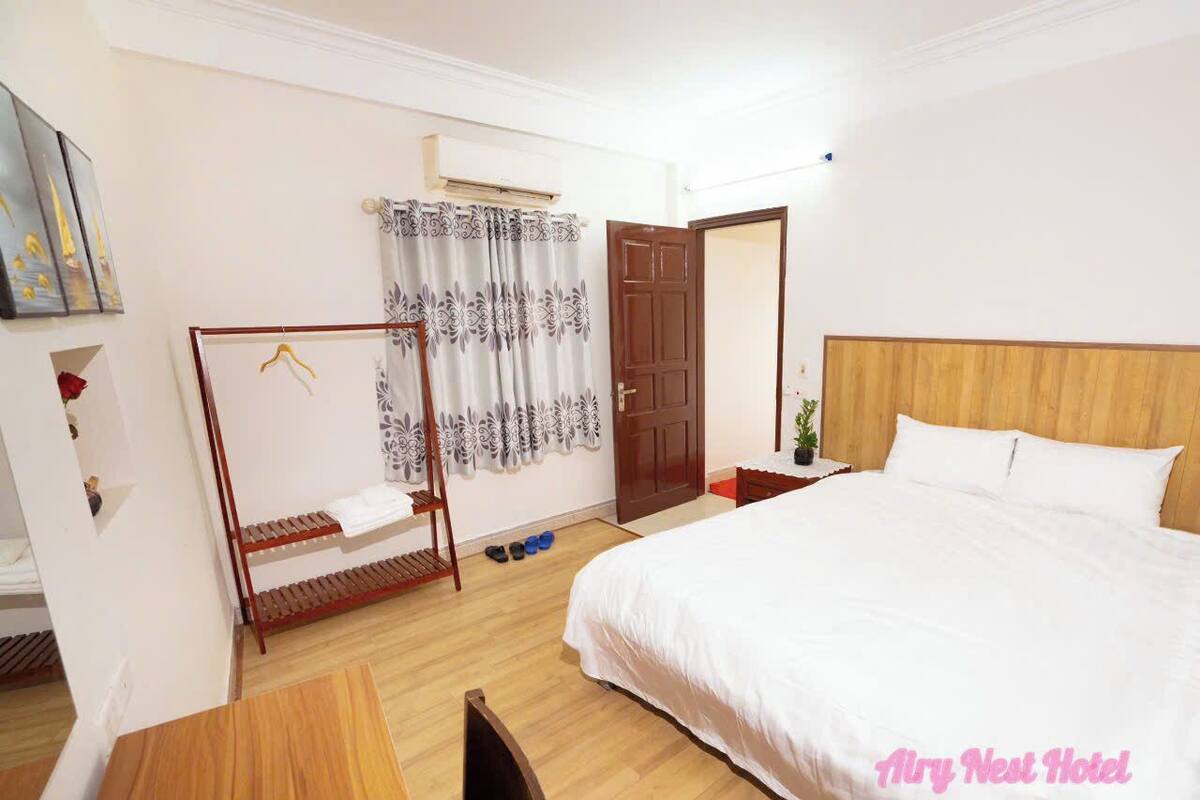 Quarto de hotel com cama de madeira, um banquinho de madeira, uma mesa pequena, um guarda-roupa, uma porta e uma janela com cortinas.