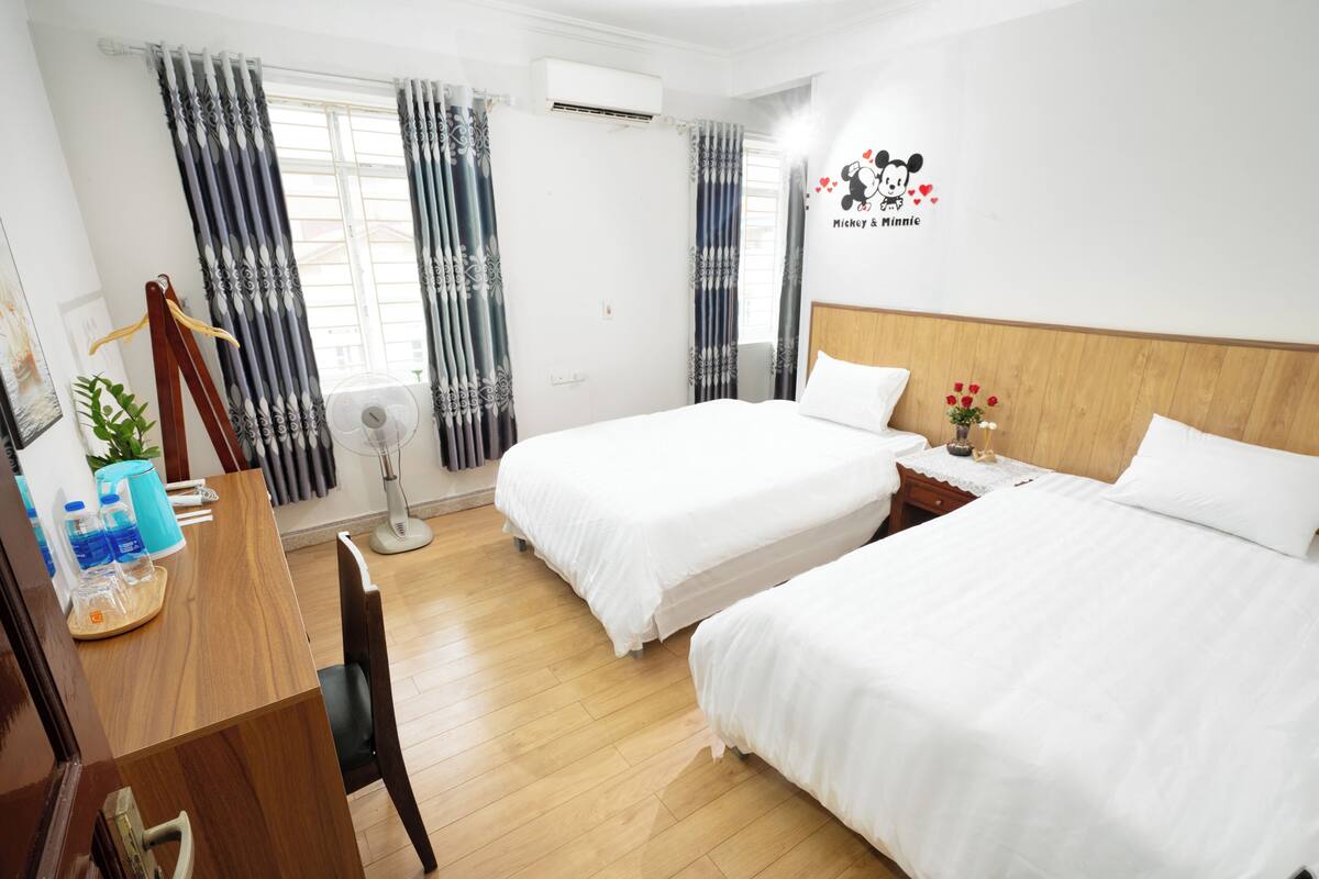 Quarto de hotel com duas camas, uma escrivaninha, uma cadeira, uma pequena mesa com um vaso de flores e um objeto decorativo na parede.