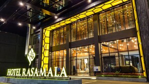Uma entrada de hotel moderna com grandes janelas de vidro, uma placa iluminada e um interior bem iluminado visível pelas janelas.