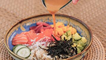 Uma tigela de sashimi com diversos acompanhamentos e uma mão despejando molho sobre ele.