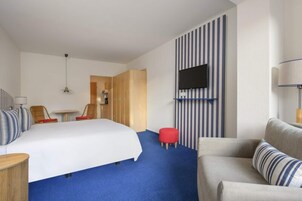 Quarto de hotel com cama, televisão na parede, um banquinho vermelho, sofá e uma mesa pequena com cadeiras.