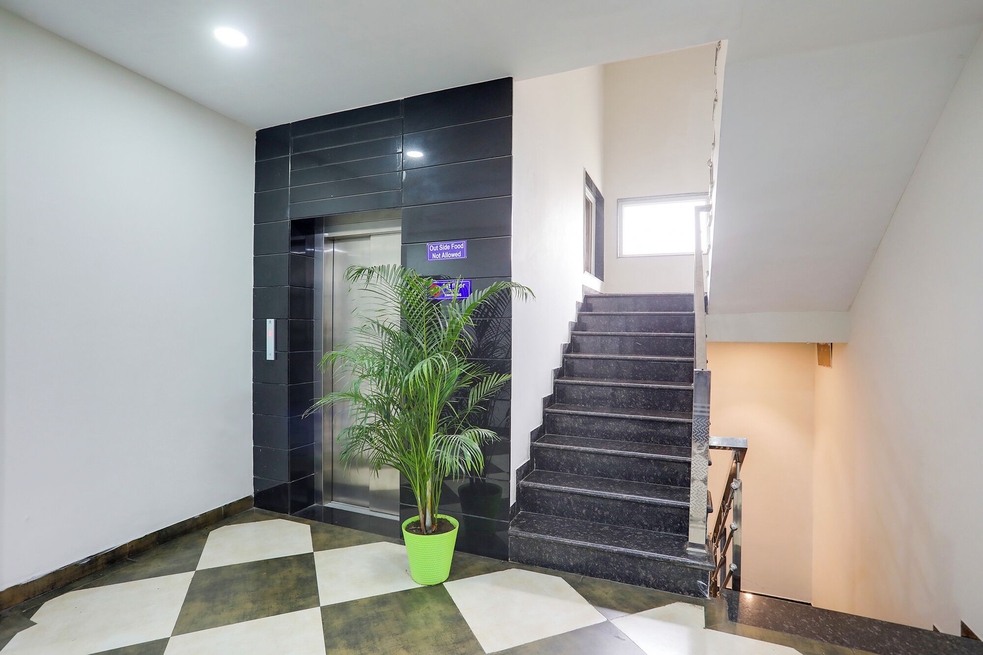 Interior moderno com piso xadrez, escada e planta em vaso perto do elevador.