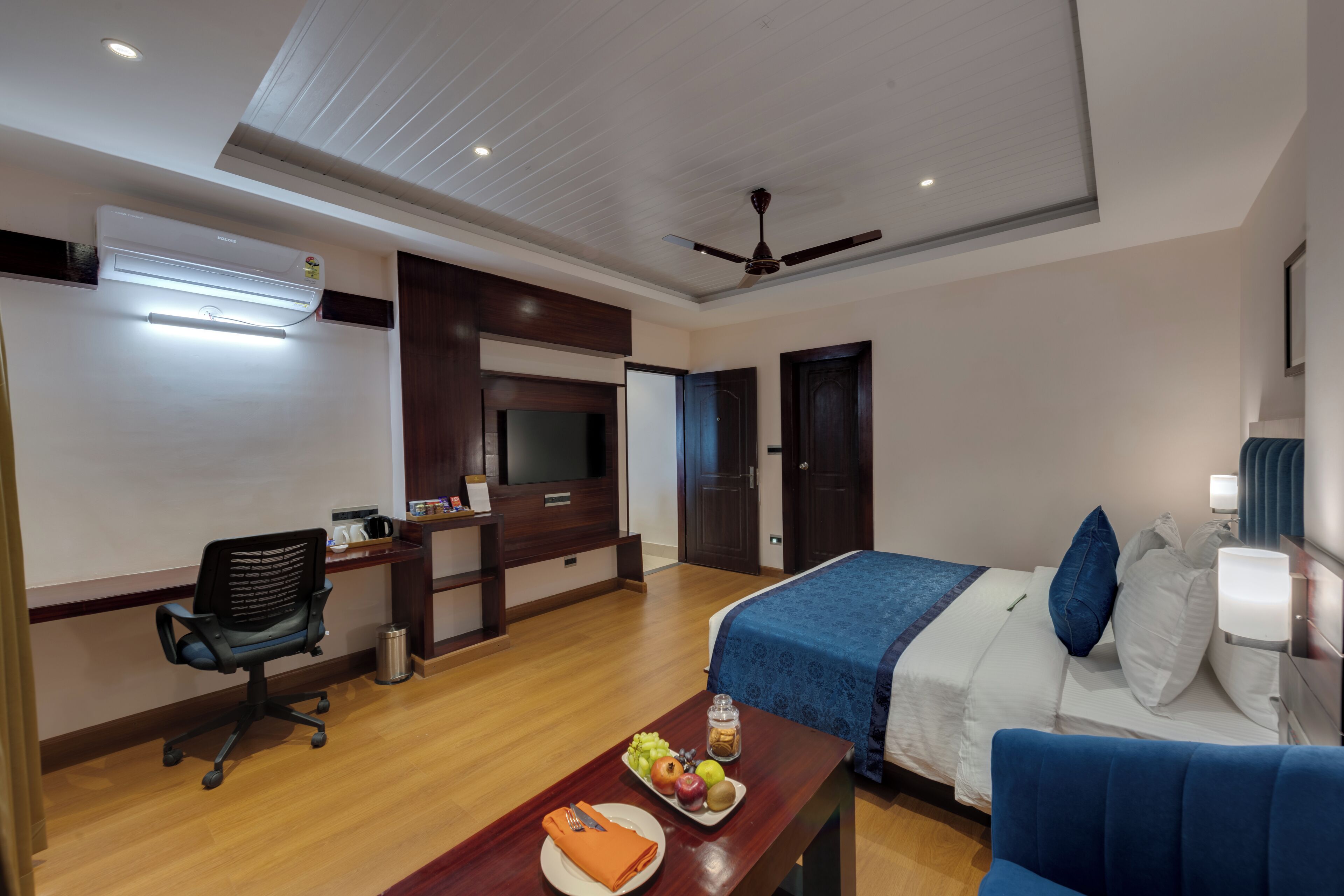Quarto de hotel com cama, secretária com cadeira, televisão e mesa de jantar com um prato de frutas.
