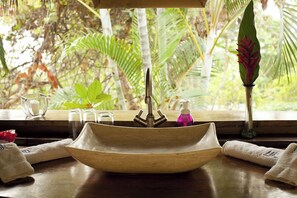 Uma bacia de madeira, uma torneira, um recipiente de vidro e um dispensador de sabonete sobre uma mesa de madeira junto a uma janela com vista para folhagem tropical.