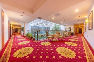 Corredor de hotel com carpete vermelho, área de refeições com cadeiras e uma mesa, e uma janela grande.