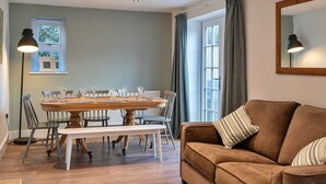 Uma área de refeições com uma mesa de madeira, cadeiras e um banco. Há um sofá marrom com almofadas listradas e um espelho na parede.