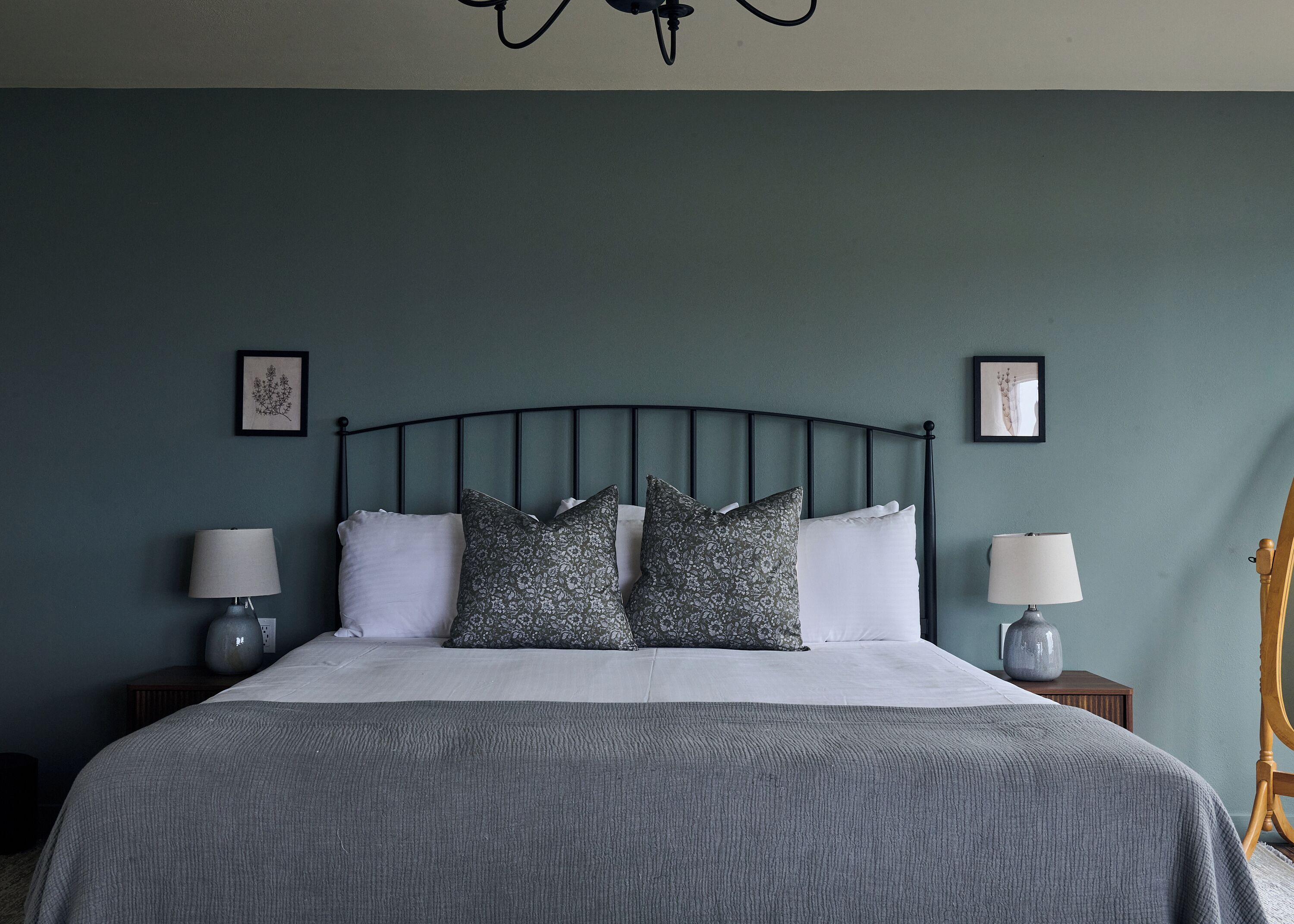 Cama bem arrumada com manta cinza e almofadas estampadas, com duas mesas de cabeceira com abajures, em frente a uma parede verde-água com obras de arte emolduradas.