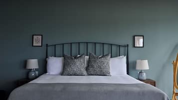 Cama bem arrumada com manta cinza e almofadas estampadas, com duas mesas de cabeceira com abajures, em frente a uma parede verde-água com obras de arte emolduradas.