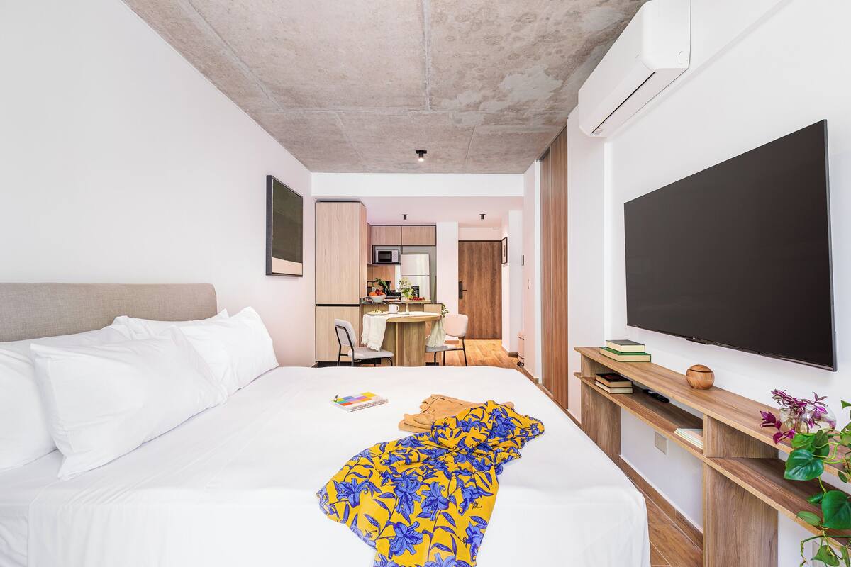Quarto de hotel com cama, televisão, escrivaninha e cozinha compacta.