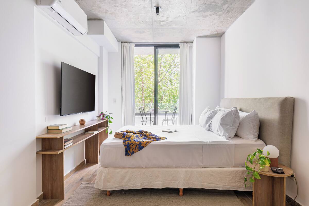 Quarto moderno com cama grande, televisão de tela plana, escrivaninha e varanda com vista para árvores.