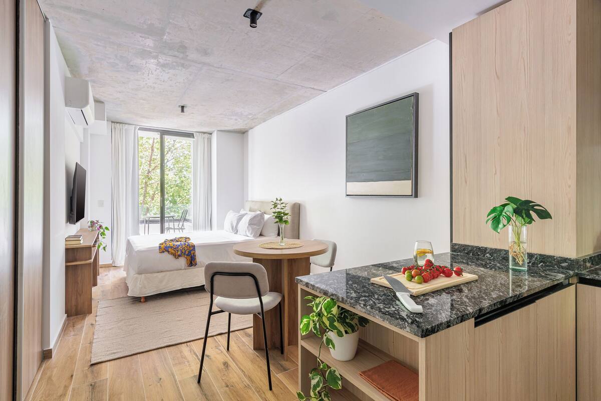 Quarto de hotel moderno com cama, área de refeições, pequena cozinha e uma grande janela.