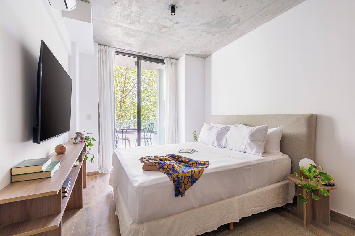 Quarto moderno com uma cama grande, televisão de tela plana, mesa de cabeceira com livros e vista para o exterior.