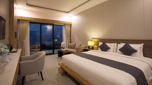 Quarto de hotel moderno com uma cama grande, uma escrivaninha com cadeira, uma mesinha e um sofá.