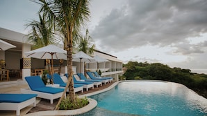 Área da piscina com espreguiçadeiras e guarda-sóis, uma palmeira e vista para a paisagem.