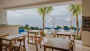 Uma área de refeições ao ar livre com mesas e cadeiras de madeira, com vista para uma praia com guarda-sóis e palmeiras.
