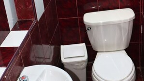 Uma casa de banho com uma pia branca, uma sanita e um contentor de lixo. As paredes estão revestidas com uma combinação de azulejos brancos e avermelhados.