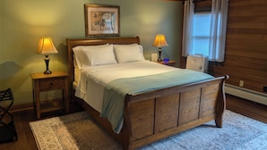 Uma cama de madeira com cabeceira, duas mesas de cabeceira com abajures, um quadro na parede e uma janela com cortinas.