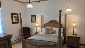 Uma cama com dossel, um painel de cabeceira, duas mesas de cabeceira com luminárias, uma cadeira e quadros nas paredes.
