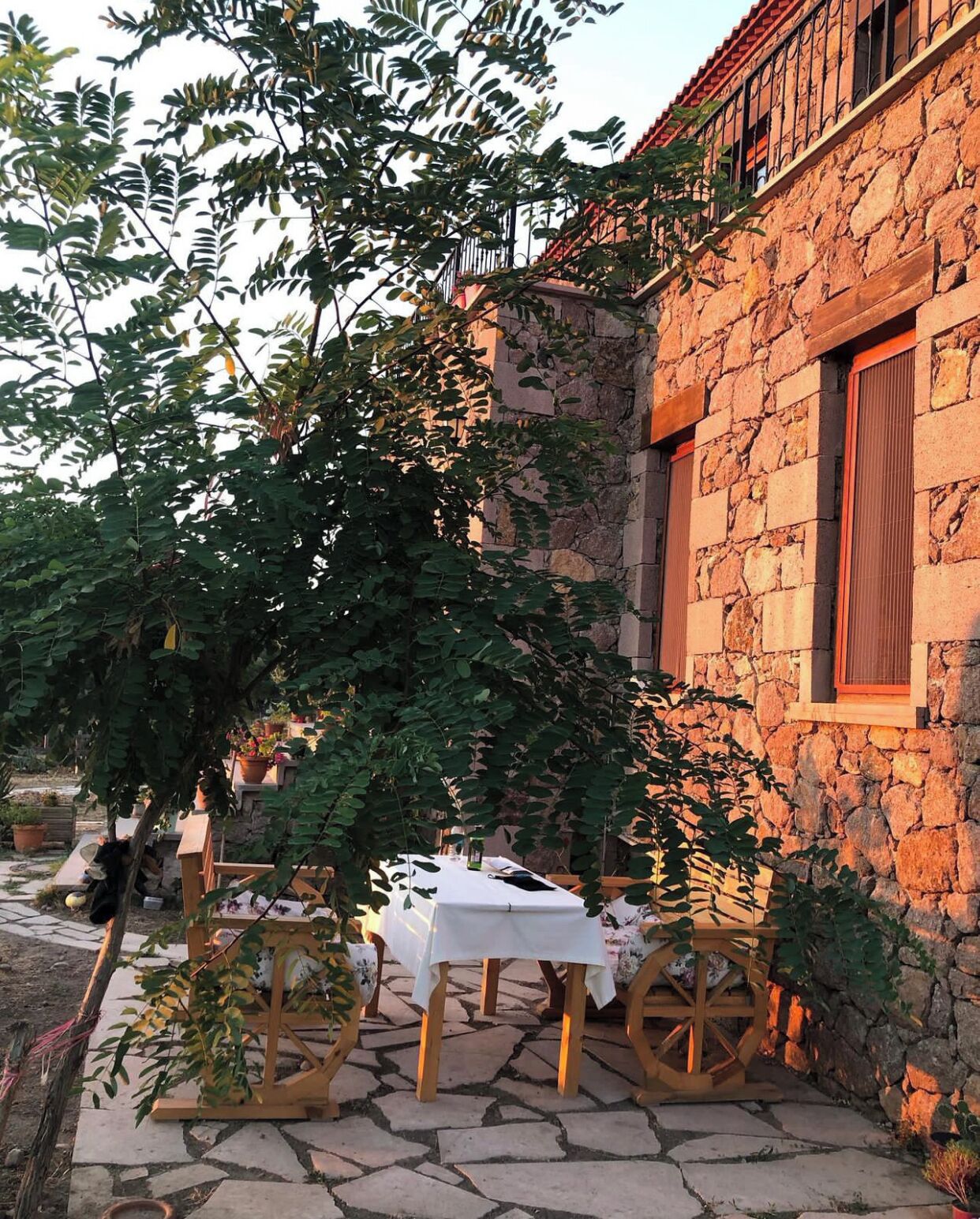 Um edifício de pedra com fachada rústica, uma mesa posta para refeição e uma árvore oferecendo sombra.