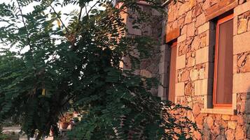 Um edifício de pedra com fachada rústica, uma mesa posta para refeição e uma árvore oferecendo sombra.