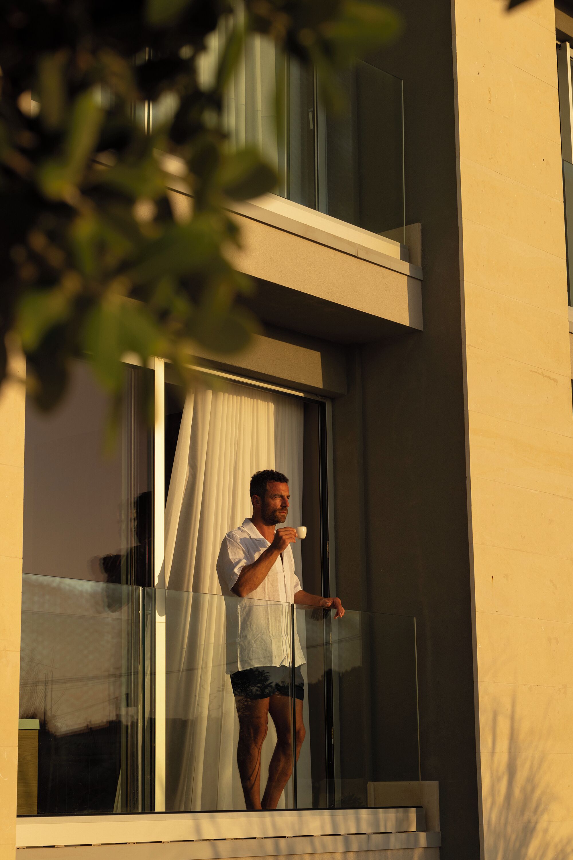 Um homem está em uma varanda com porta de vidro e cortinas brancas.