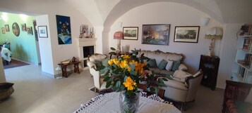 Uma sala de estar com lareira, um sofá, uma mesa de centro com um vaso de flores e uma estante de livros.
