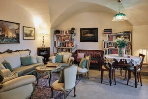 Uma sala de estar ampla com sofá, cadeiras, mesa de centro e mesa de jantar com cadeiras. Estantes repletas de livros e objetos decorativos.