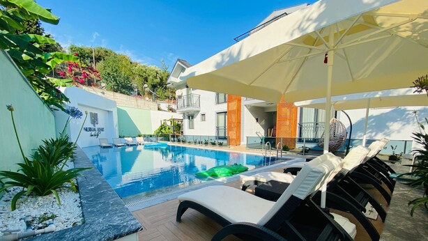 Área de piscina com espreguiçadeiras e guarda-sóis, cercada por vegetação e um edifício com varanda.