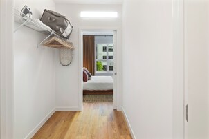 Um corredor estreito com piso de madeira, uma porta branca que dá para um quarto com cama e uma janela com cortinas.