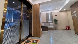 Um saguão de hotel com balcão de recepção, área de estar com sofá e mesa de centro, e um extintor de incêndio na parede.