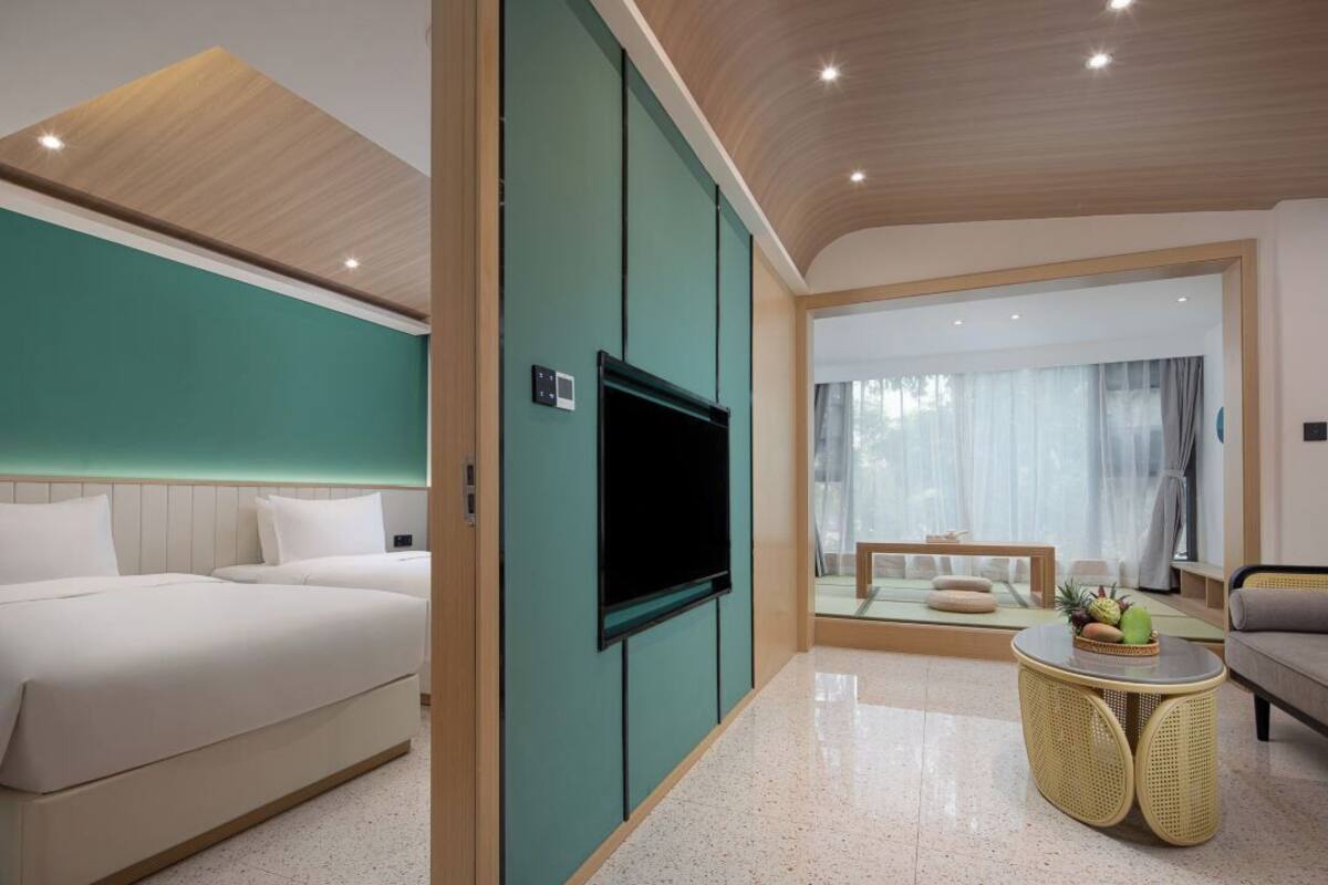 Quarto de hotel com uma cama, televisão, uma mesinha redonda com frutas e vista para o exterior através de uma porta de vidro.
