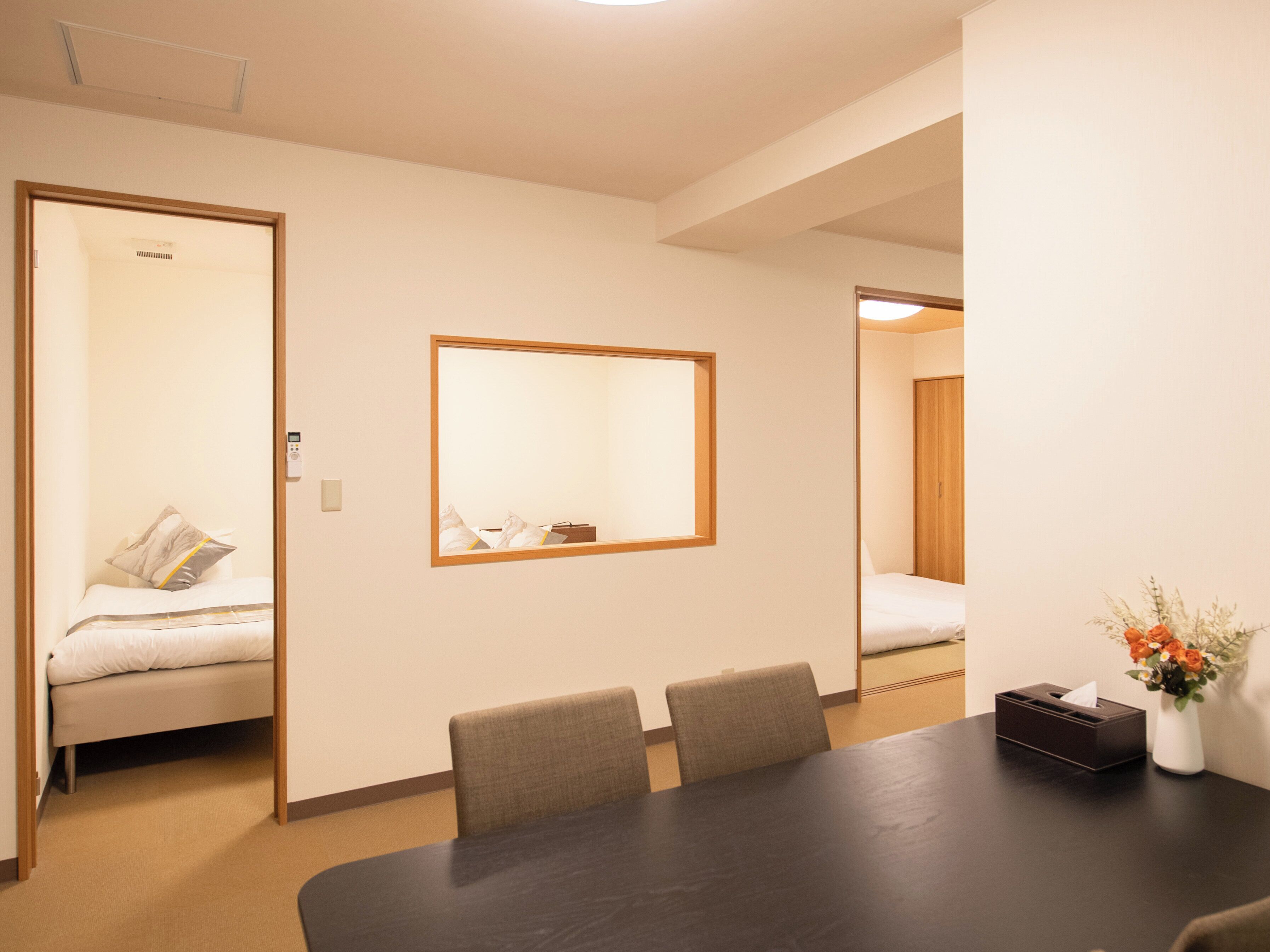 Quarto de hotel com cama, mesa de cabeceira, cadeira, mesa com cadeiras, espelho e um vaso com flores.