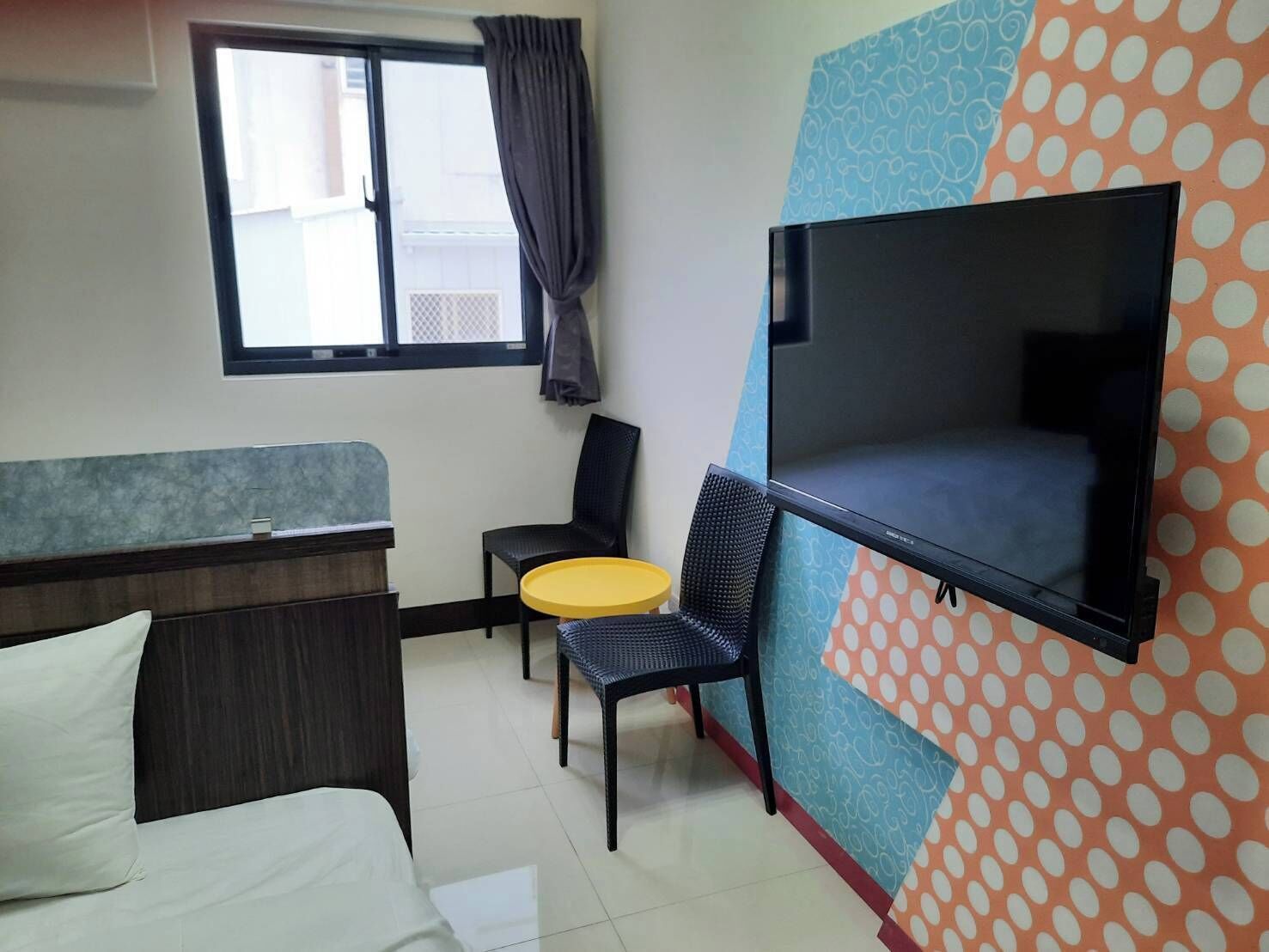 Quarto de hotel com cama, uma pequena mesa com duas cadeiras, TV fixada na parede e janela com cortinas.