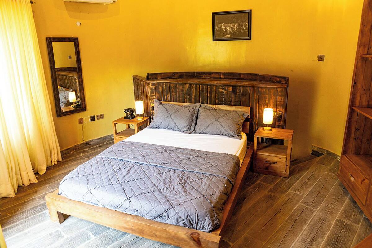 Uma cama com um cobertor acolchoado, duas mesas de cabeceira com luminárias, uma cabeceira de madeira, um espelho e um quadro na parede.