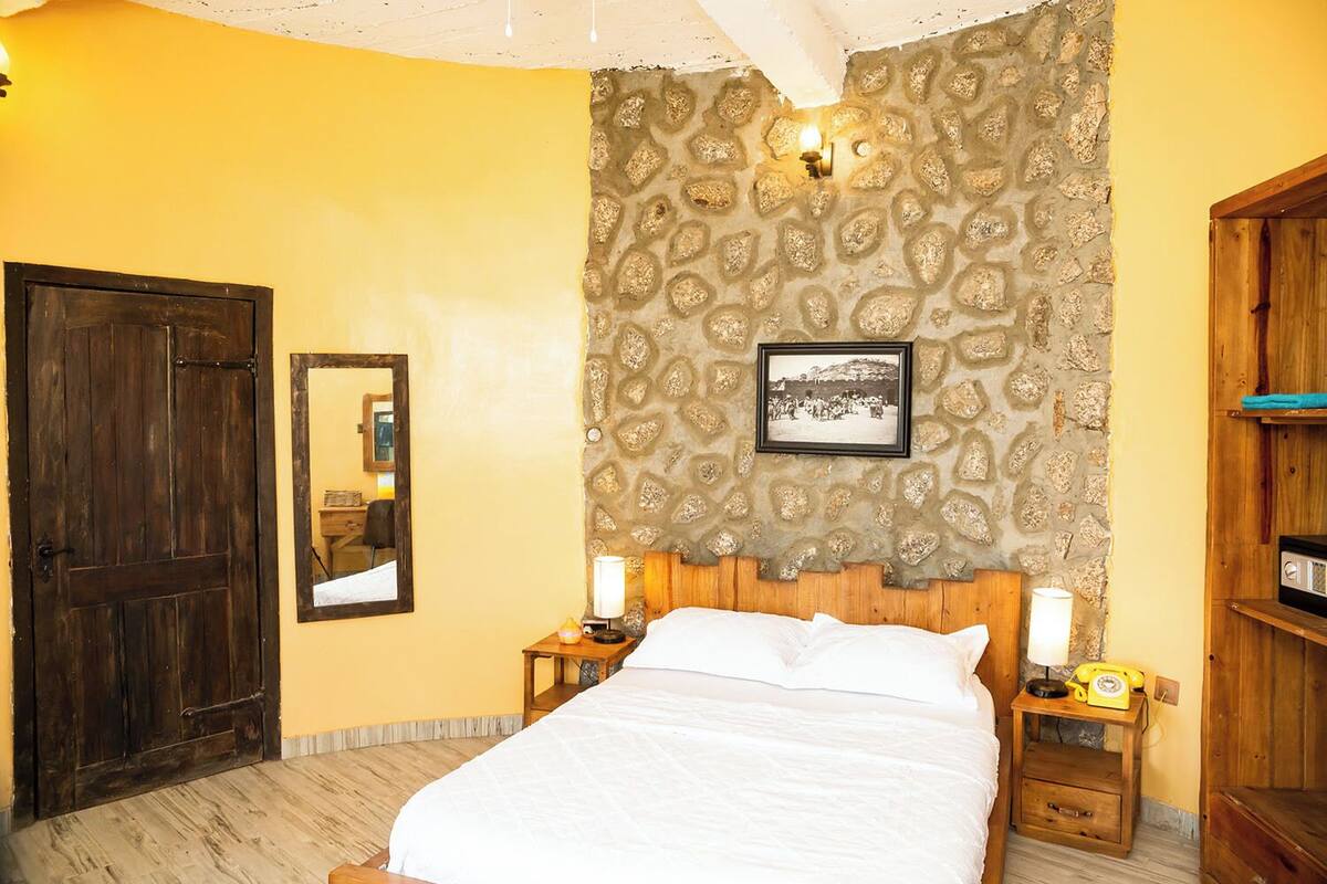 Quarto com porta de madeira, cama com roupa de cama branca, duas mesas de cabeceira com abajures, um espelho e parede com padrão de pedra.