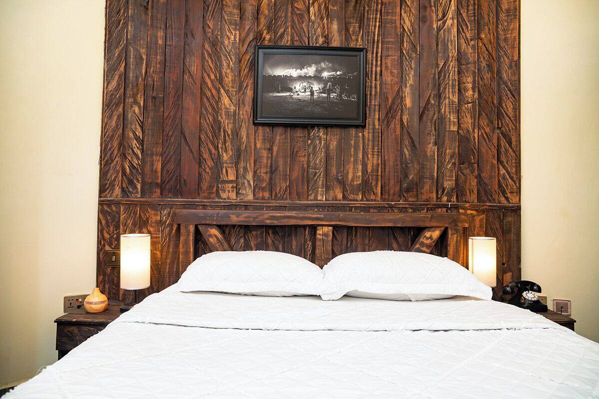 Uma cama com roupa de cama branca, cabeceira de madeira, um quadro emoldurado, duas luminárias de cabeceira e um telefone antigo.