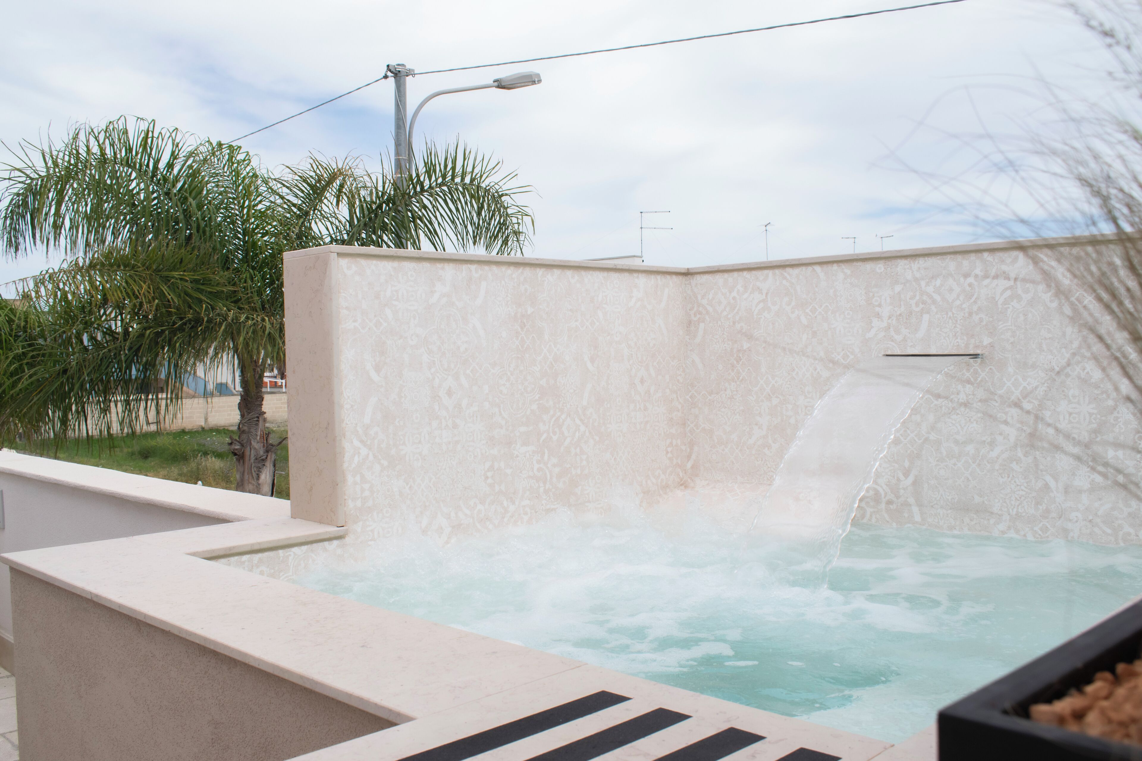 Uma banheira de hidromassagem com cascata, rodeada por um muro bege e palmeiras.