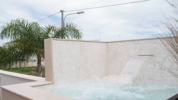 Uma banheira de hidromassagem com cascata, rodeada por um muro bege e palmeiras.