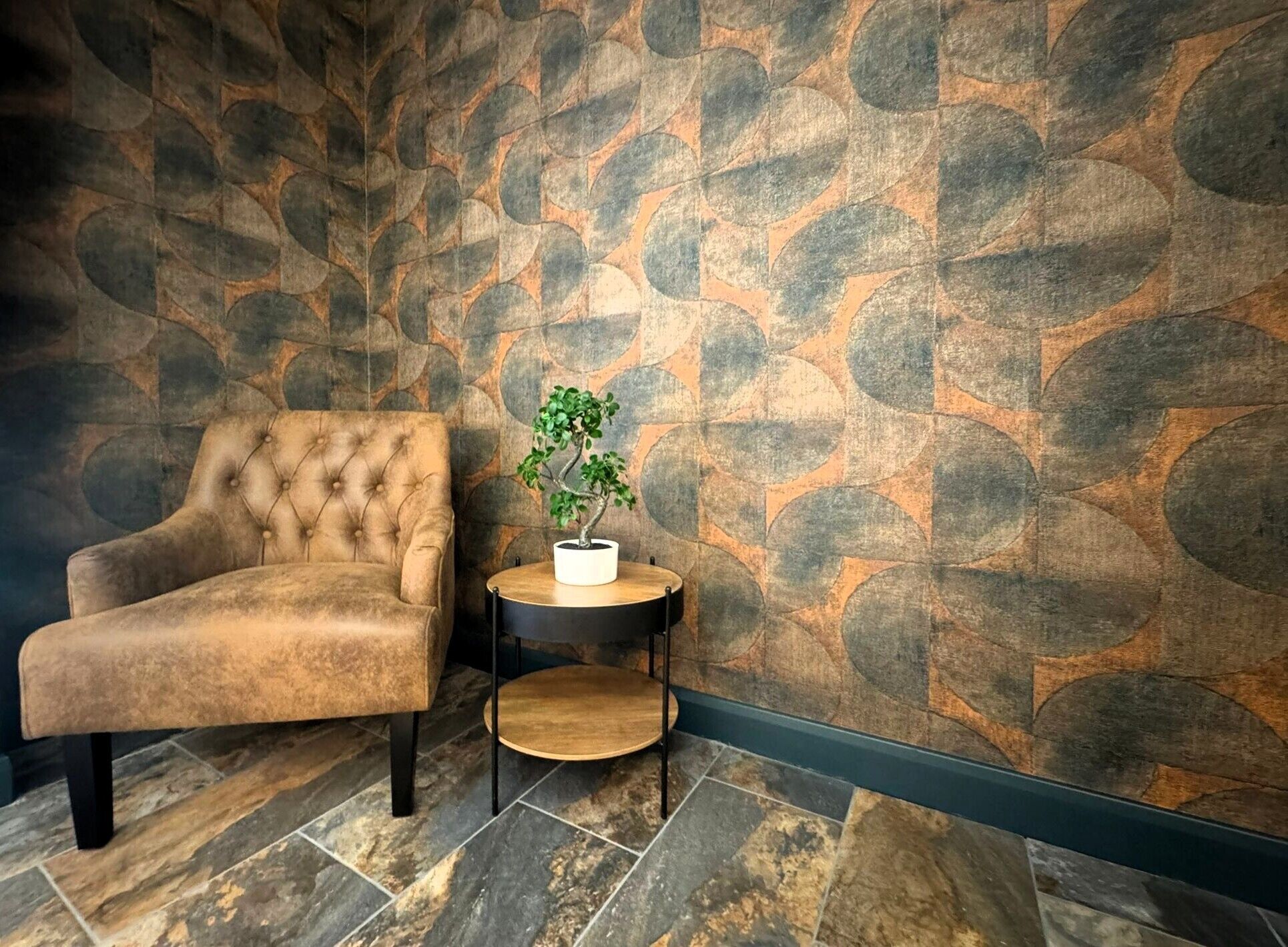 Uma poltrona com detalhes em tufos e uma mesa de madeira redonda com uma planta em vaso, dispostas diante de uma parede com textura que imita pedra.