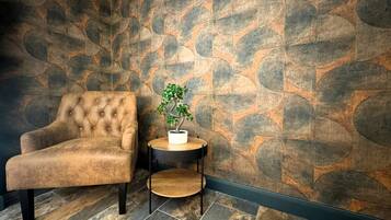 Uma poltrona com detalhes em tufos e uma mesa de madeira redonda com uma planta em vaso, dispostas diante de uma parede com textura que imita pedra.