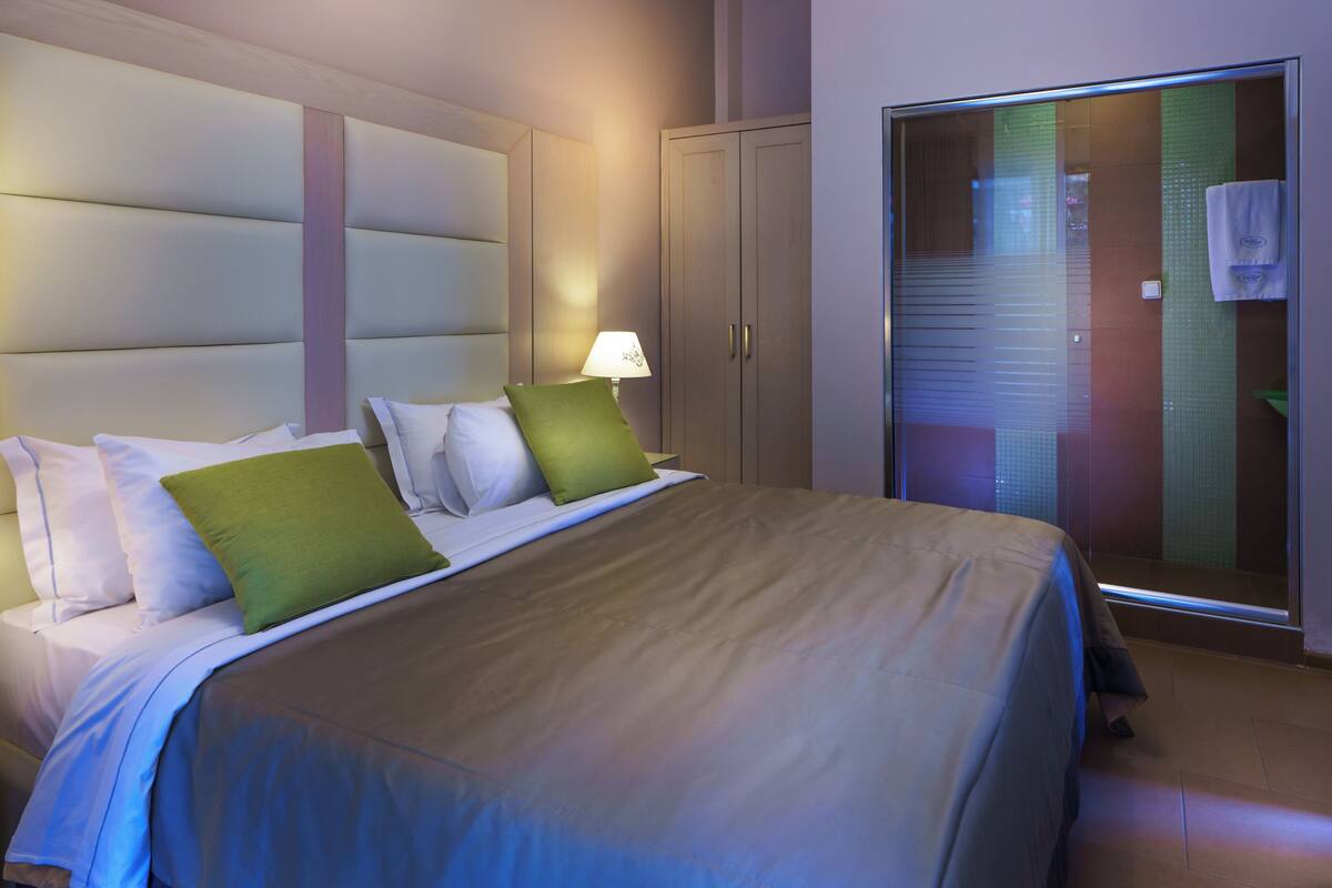 Quarto de hotel com uma cama grande, mesa de cabeceira com abajur, guarda-roupa e porta de vidro deslizante para um banheiro.