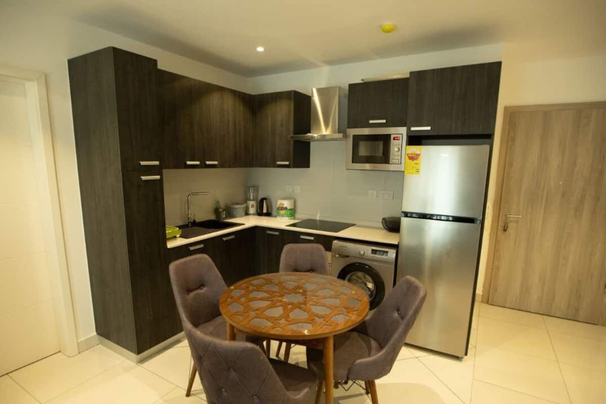 Uma cozinha compacta com armários escuros, micro-ondas, frigorífico, máquina de lavar e uma mesa redonda com quatro cadeiras.