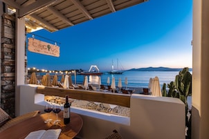 Terraço à beira-mar com vista para o mar, um barco e uma ponte. Há espreguiçadeiras e guarda-sóis, além de uma mesa com uma garrafa de vinho e copos.