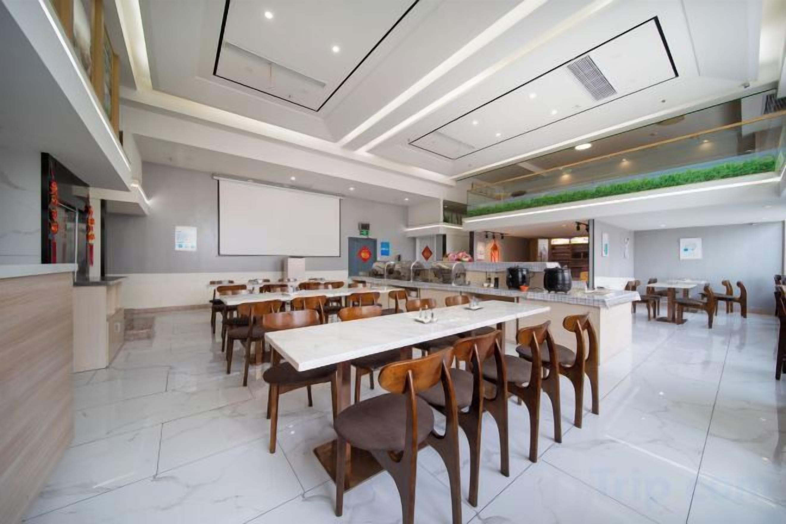 Uma sala de conferências ampla com uma mesa comprida, várias cadeiras, uma tela de projetor e uma área de cozinha ao fundo.