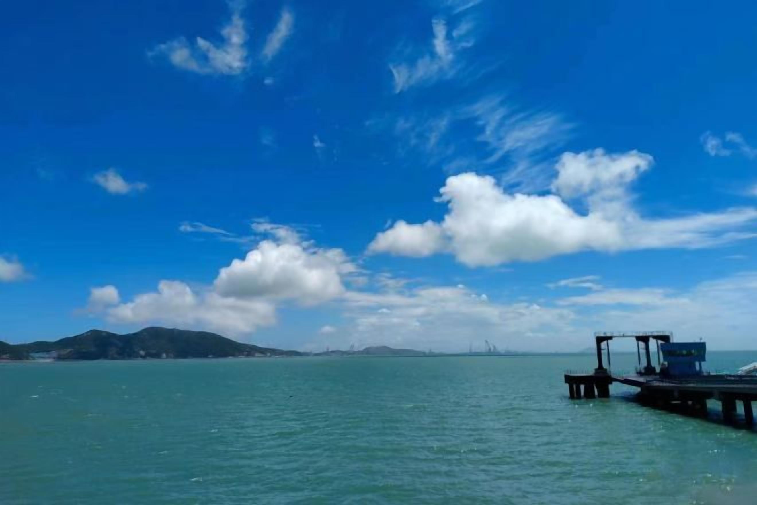 Céu azul e límpido, mar calmo e um cais que se projeta sobre a água.