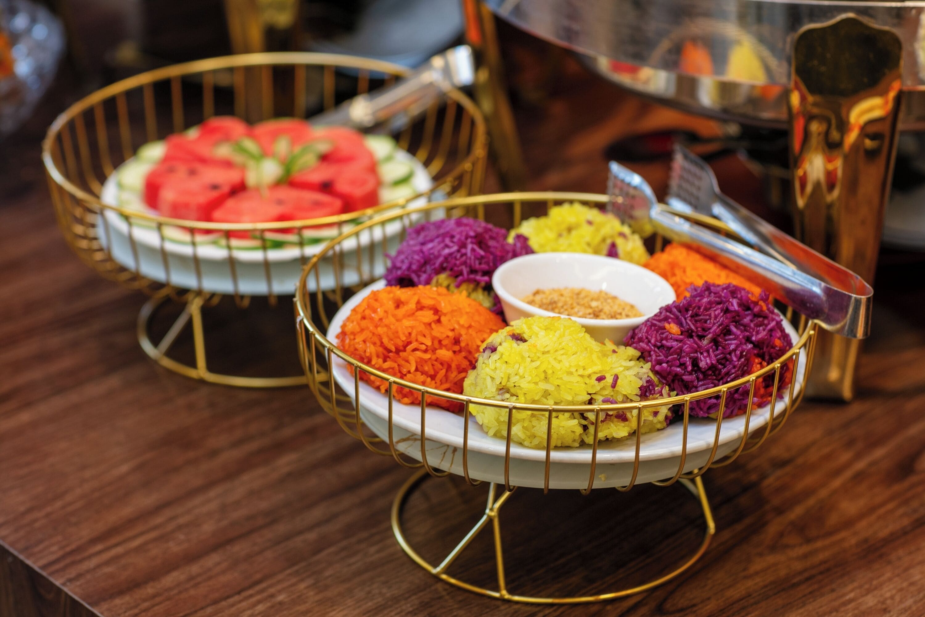 Duas tigelas douradas sobre uma mesa de madeira, cada uma com pratos de arroz coloridos e uma pequena tigela de especiarias.