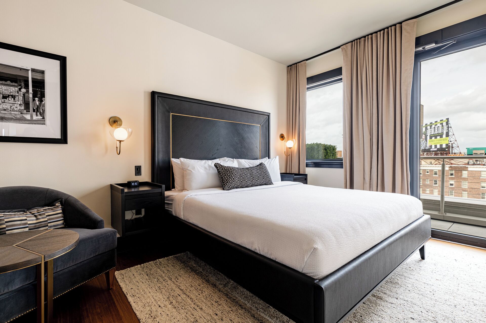 Quarto de hotel moderno com uma cama grande, uma mesa de apoio, uma cadeira e vista da cidade através de amplas janelas.