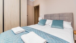 Cama bem arrumada com colcha azul e almofadas brancas, um guarda-roupa de madeira e uma porta de madeira ao fundo.